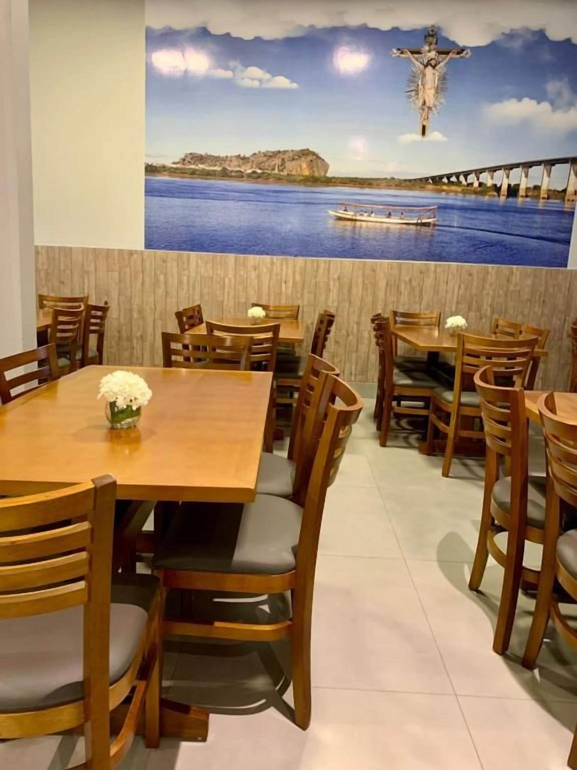 Interior de restaurante com mesas e cadeiras de madeira, um mural com paisagem costeira e vista para um corpo de água.