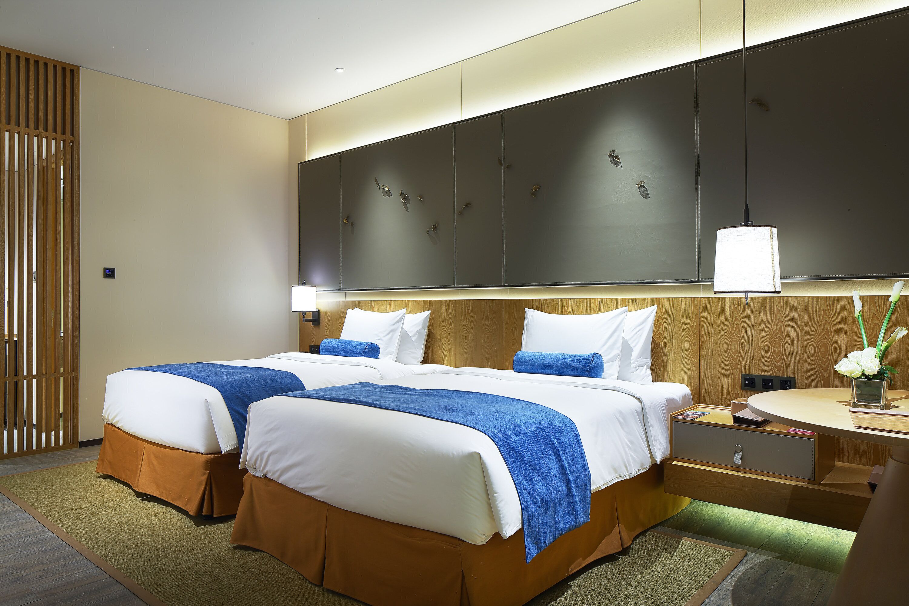 Quarto de hotel com duas camas, cabeceira de madeira, mesa de cabeceira com abajur e um pequeno vaso com flores.