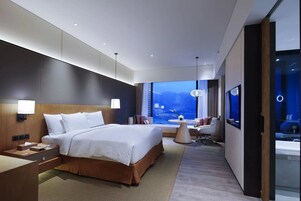 Quarto de hotel moderno com uma cama grande, escrivaninha, cadeira e vista para a paisagem urbana.