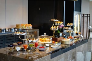 Buffet com diversos alimentos e bebidas, incluindo uma mesa de sobremesas em três níveis, potes de frutas e um canto com café.