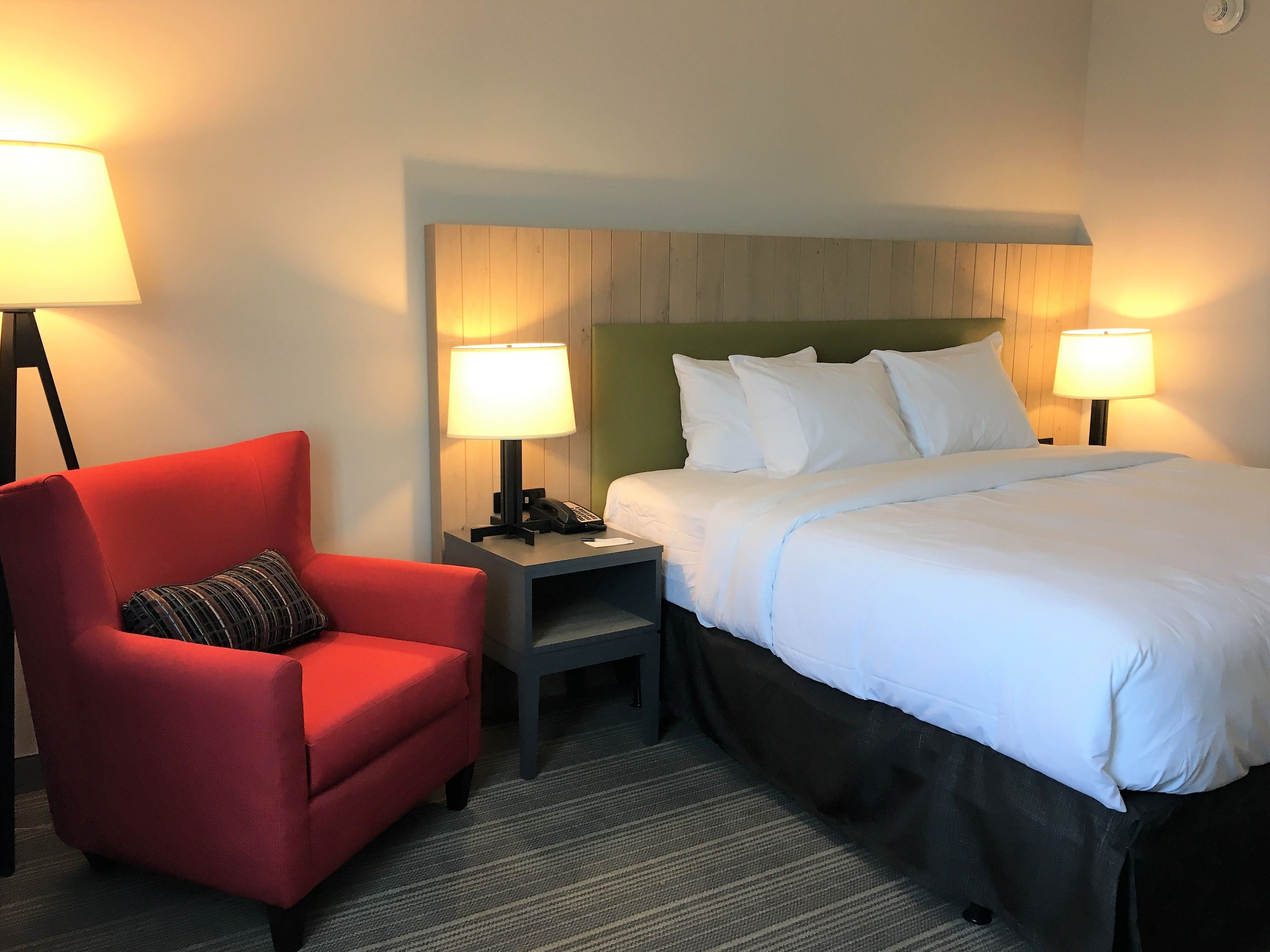Quarto de hotel com uma cama de solteiro, uma poltrona vermelha, duas mesas de cabeceira com luminárias e uma pequena mesinha de apoio.