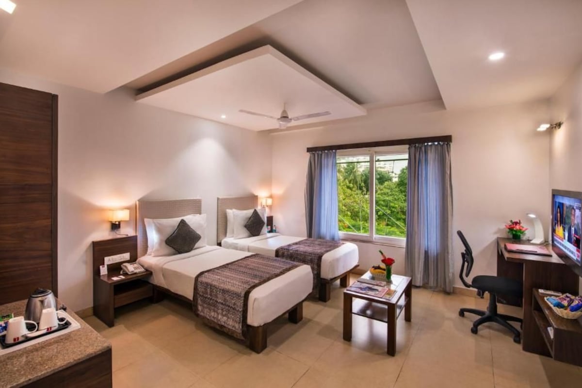 Quarto de hotel com duas camas, uma escrivaninha, uma cadeira, uma televisão e uma janela com cortinas.
