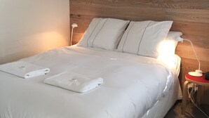 Cama bem arrumada com lençóis brancos e duas almofadas, mesa de cabeceira com abajur e cabeceira de madeira.