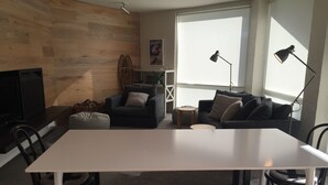 Uma sala de estar moderna com uma mesa branca, sofás escuros e uma televisão.