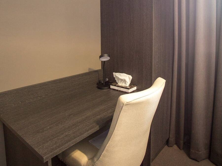 Quarto de hotel com escrivaninha de madeira escura, uma poltrona estofada de branco, luminária de mesa e caixa de lenços.