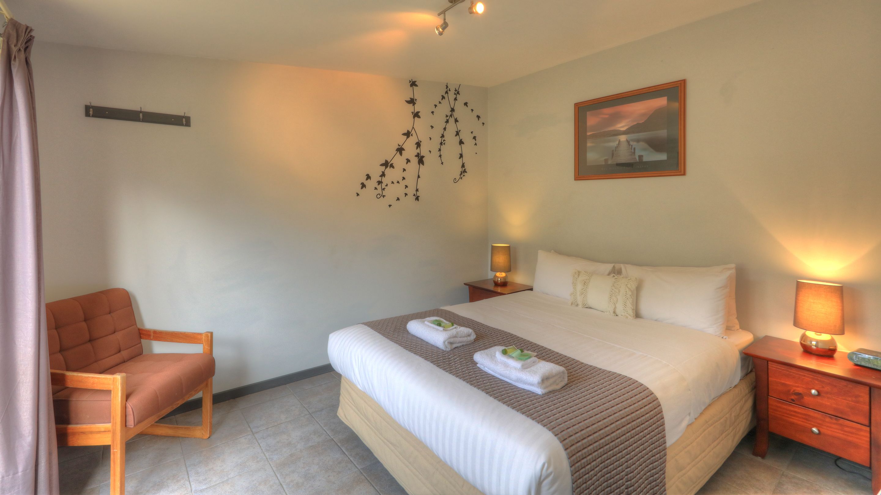 Quarto de hotel bem arrumado com uma cama grande, uma cadeira de madeira, uma mesa de cabeceira com abajur e um quadro na parede.