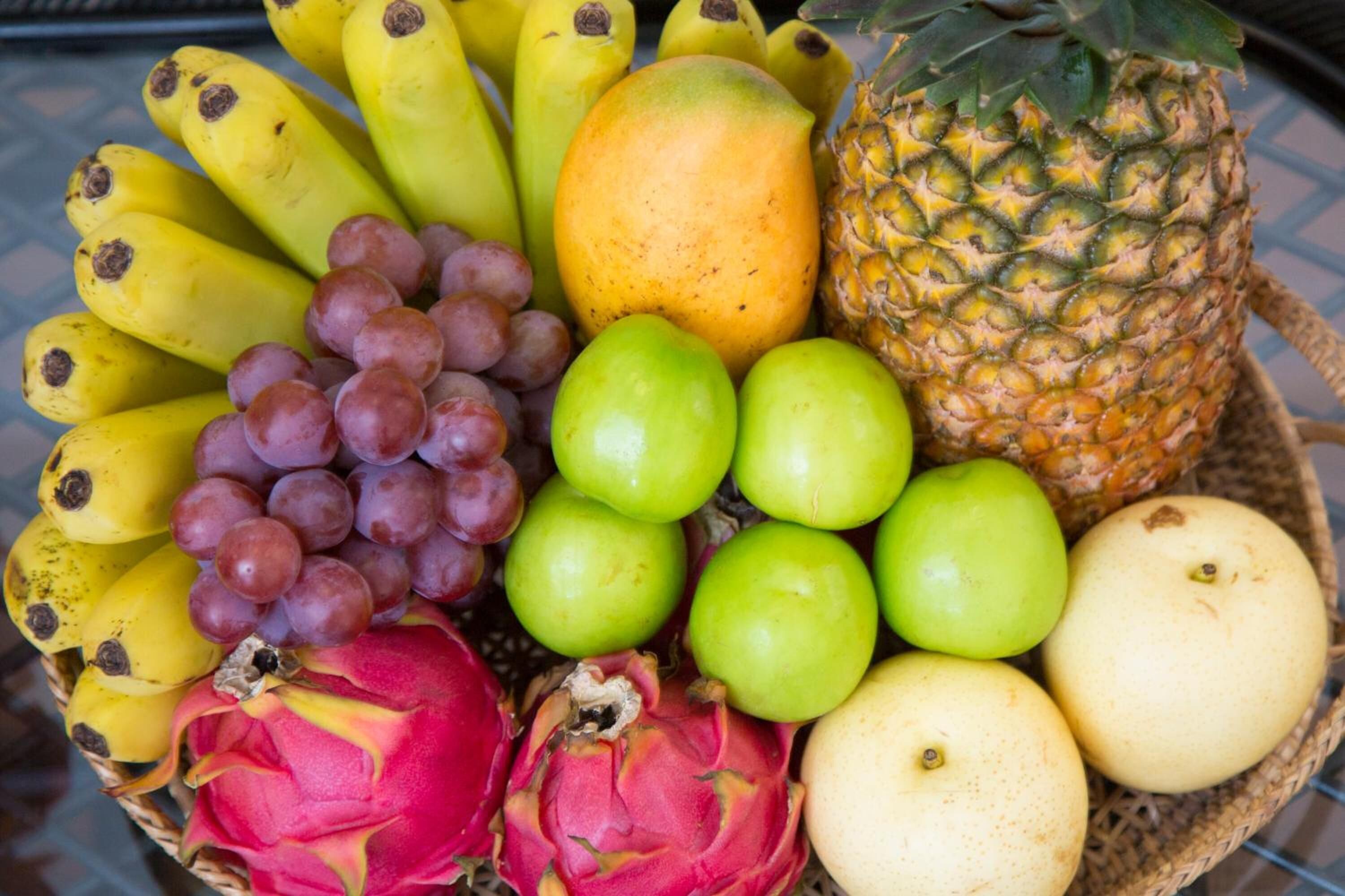 Uma cesta com diversas frutas, incluindo bananas, uvas, maçãs, pitaya, manga e abacaxi.