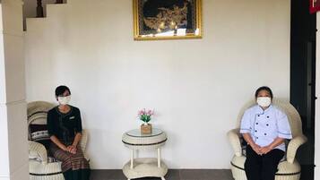 Duas pessoas sentadas em cadeiras de vime num espaço com chão de azulejos, uma mesa redonda pequena com um vaso de flores e um quadro na parede.