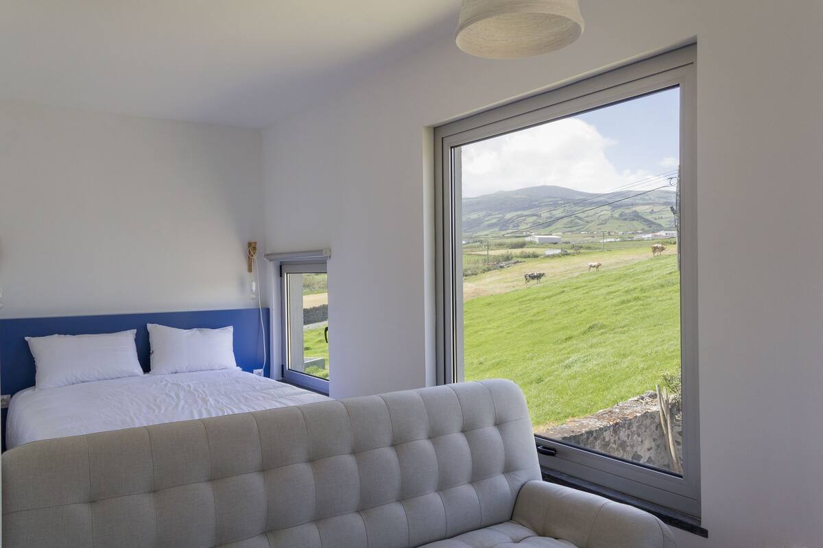 Quarto com uma janela grande, uma cama e vista para um campo verde com vacas.