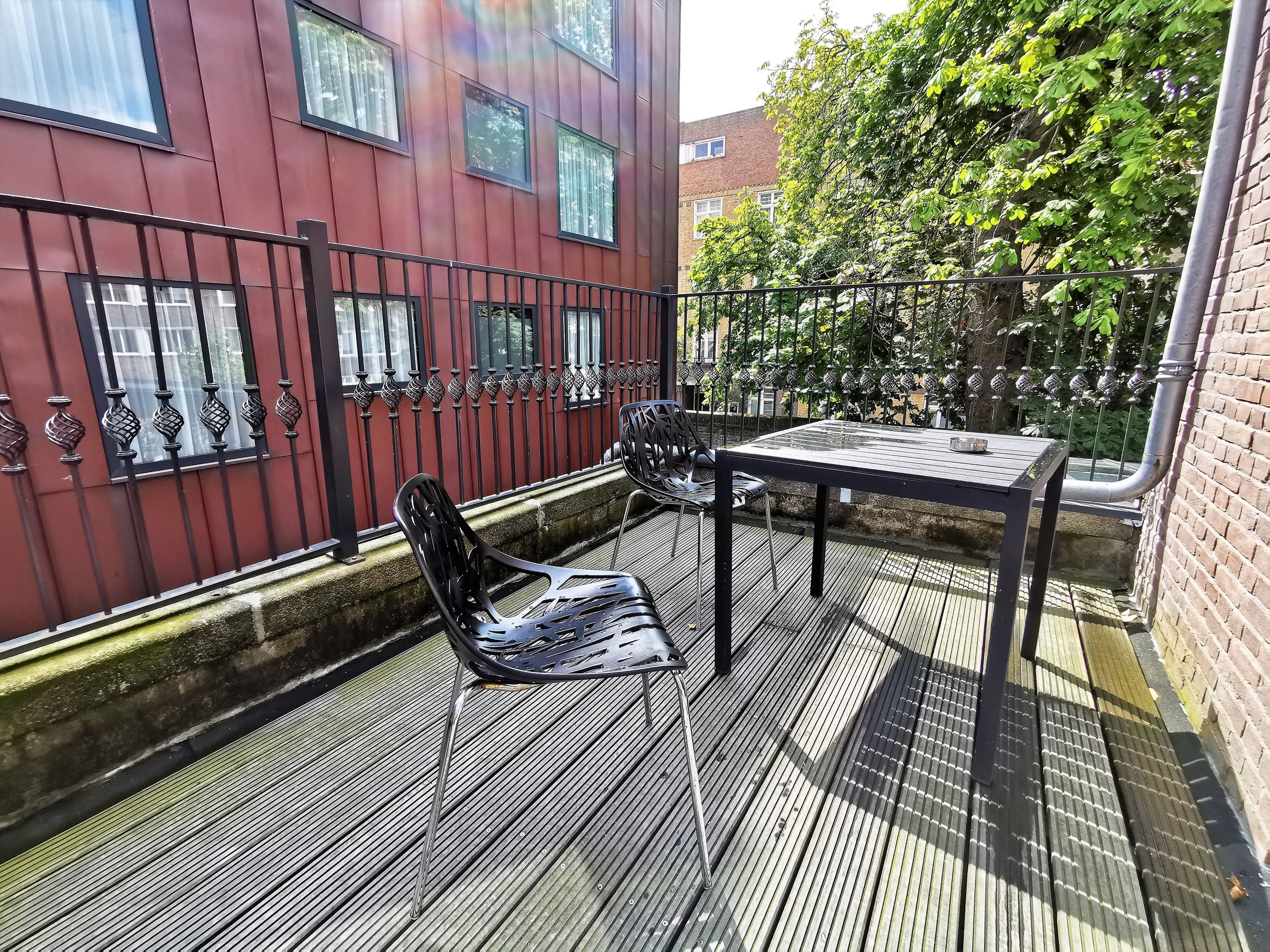 Um pátio externo com mesa e cadeiras, um edifício vermelho com janelas e uma cerca de metal.