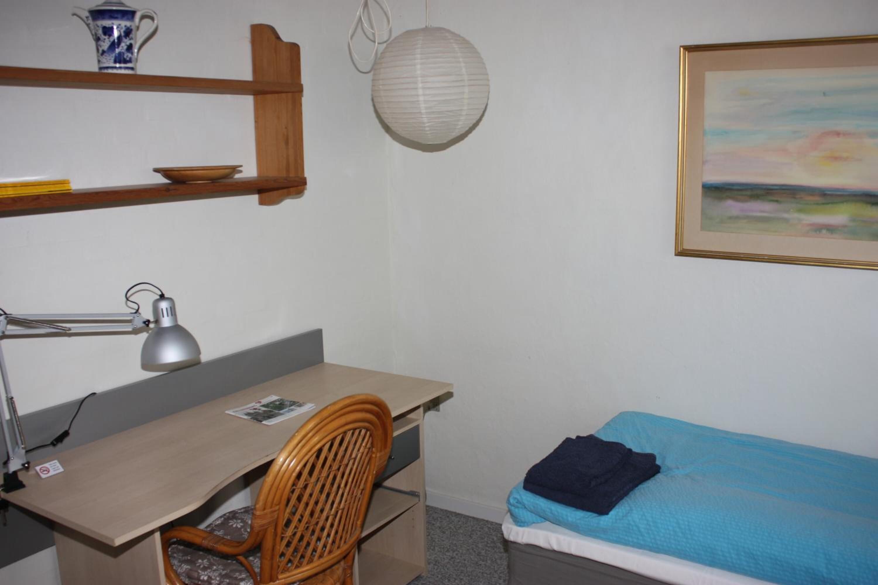 Quarto pequeno com escrivaninha, cadeira e uma cama de solteiro.