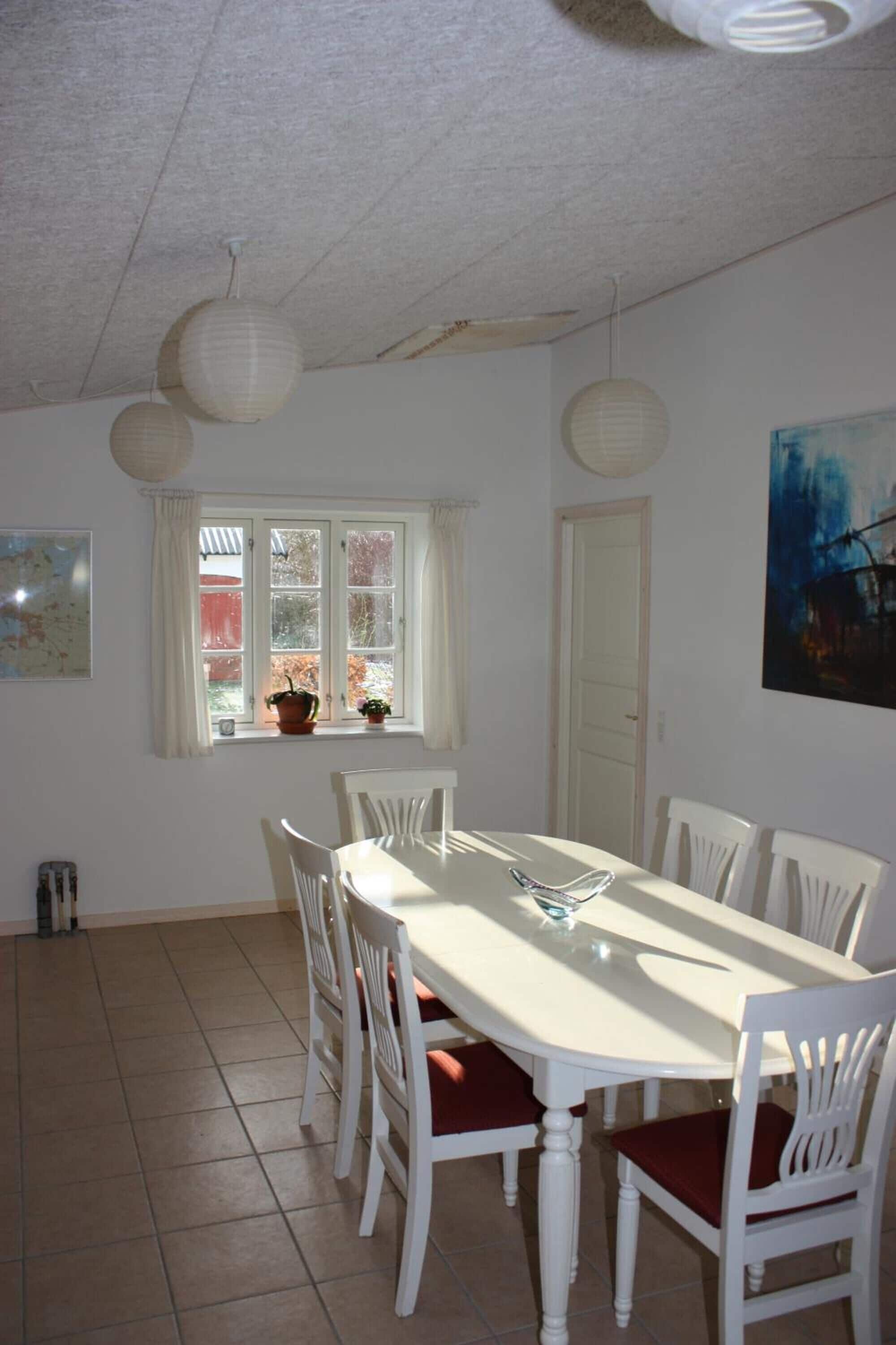 Uma área de refeições com uma mesa e cadeiras brancas, uma janela com cortinas e um quadro na parede.