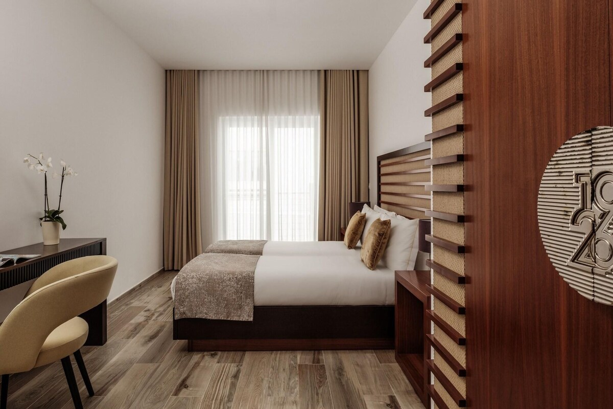 Quarto de hotel com cama, escrivaninha, cadeira, planta e uma grande janela com cortinas.