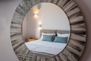 Espelho redondo com moldura de madeira refletindo um quarto com cama, almofadas e mesa de cabeceira.