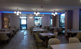 Uma sala de jantar moderna com várias mesas e cadeiras, luminárias no teto e grandes janelas.