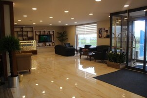 Um hall amplo com piso de mármore polido, área de estar com sofás, televisão e uma grande janela com persianas.