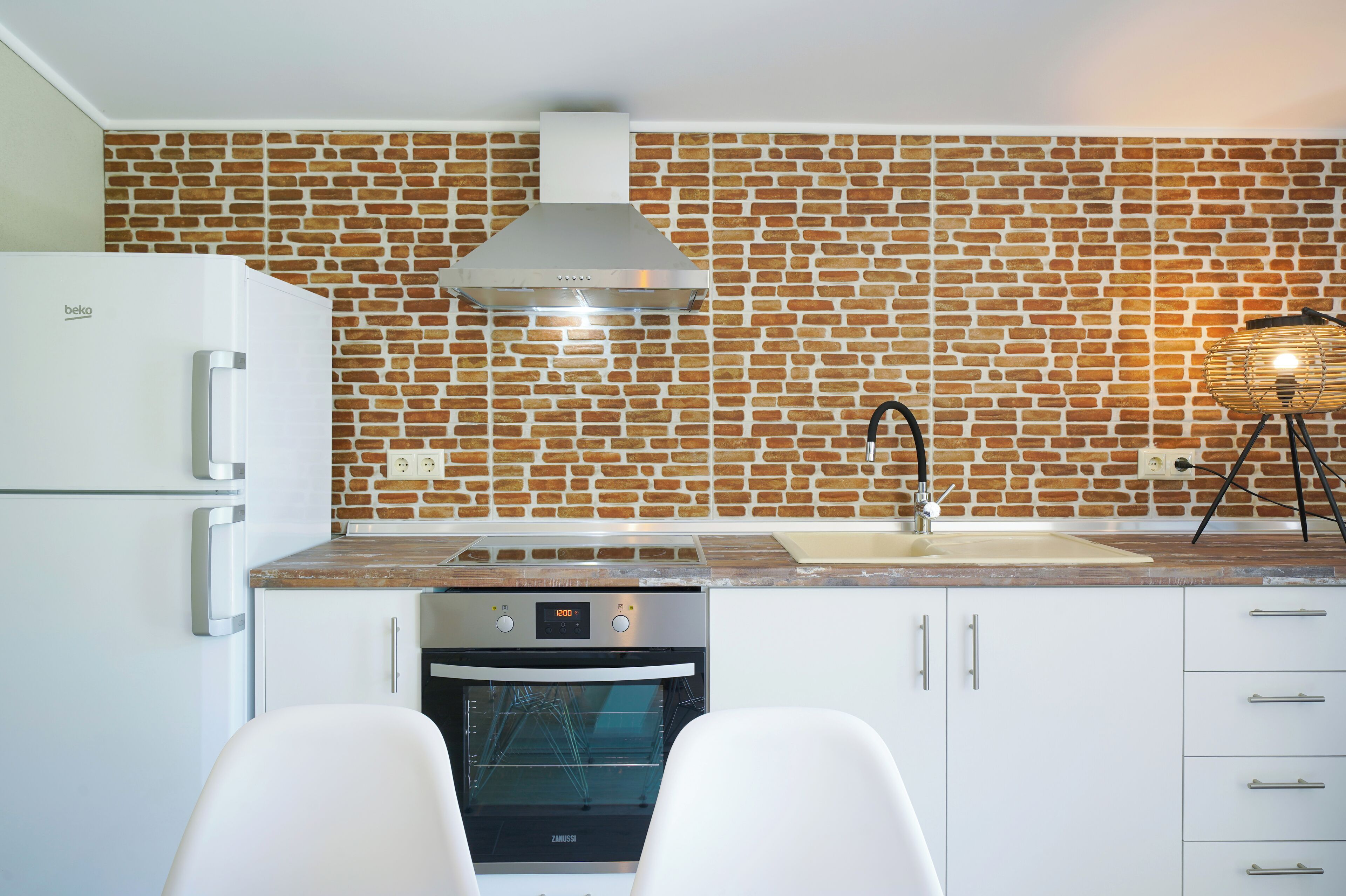 Uma cozinha com painel de parede de tijolos, coifa de aço inoxidável, forno embutido e pia com torneira moderna.