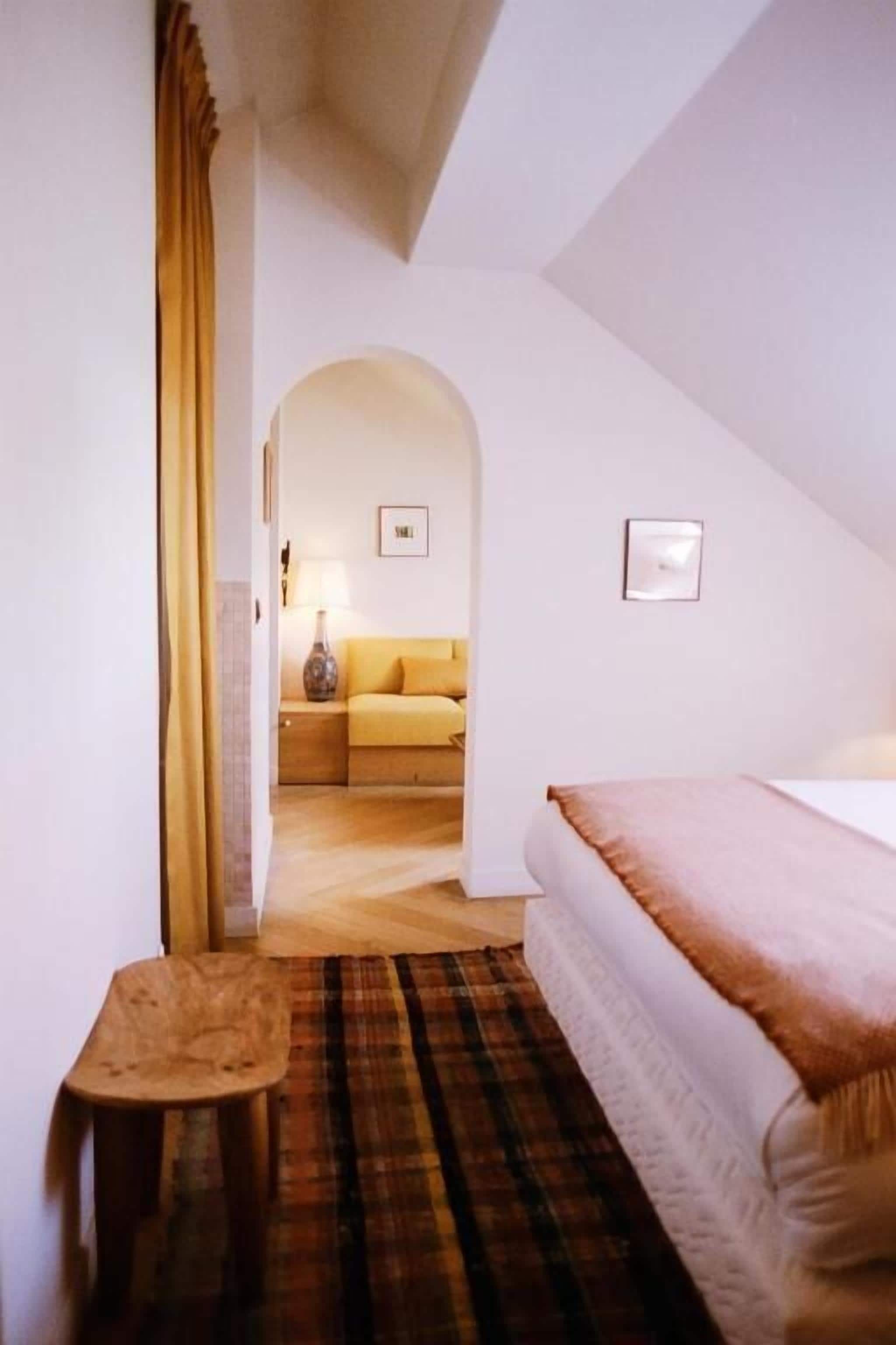 Um quarto com uma cama, um banquinho pequeno, uma luminária e um quadro pendurado na parede.