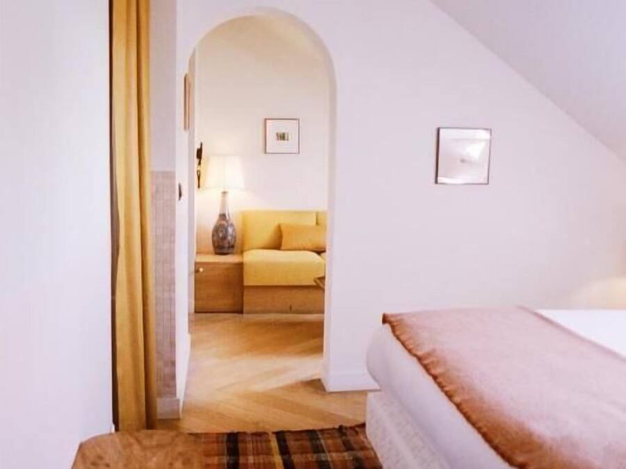 Um quarto com uma cama, um banquinho pequeno, uma luminária e um quadro pendurado na parede.