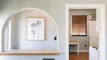 Quarto com um banco de madeira, um quadro com palmeiras e uma janela com persianas, além de uma mesa branca.