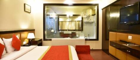 Quarto de hotel com uma cama grande, televisão, escrivaninha e banheiro visível através de um painel de vidro.