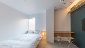 Quarto moderno com cama, mesa de cabeceira, luminária na parede e um pequeno espaço de estar com bancos.