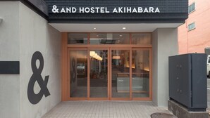Exterior do And Hostel Akihabara, com fachada moderna, portas de vidro grandes e uma placa chamativa.