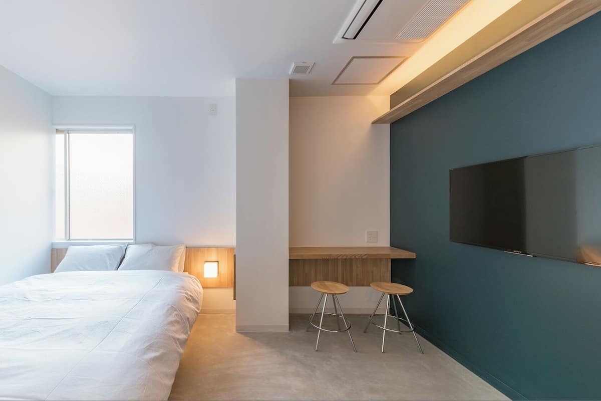 Quarto de hotel moderno com cama, escrivaninha com dois banquetes, televisão presa à parede e janela que oferece luz natural.