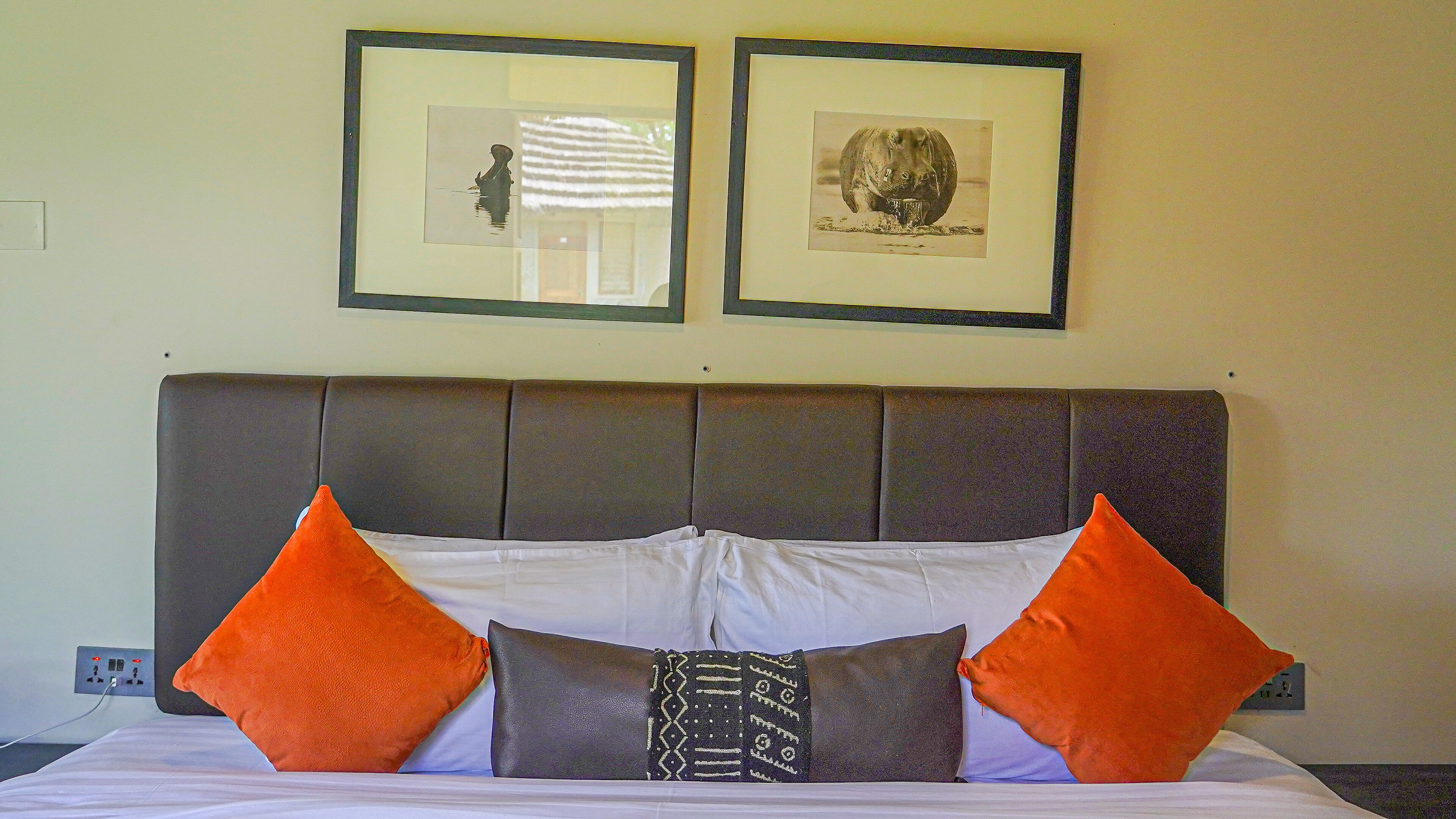 Cama com roupa de cama branca, dois travesseiros laranja e um preto, e cabeceira escura. Duas fotos emolduradas estão penduradas acima da cama.