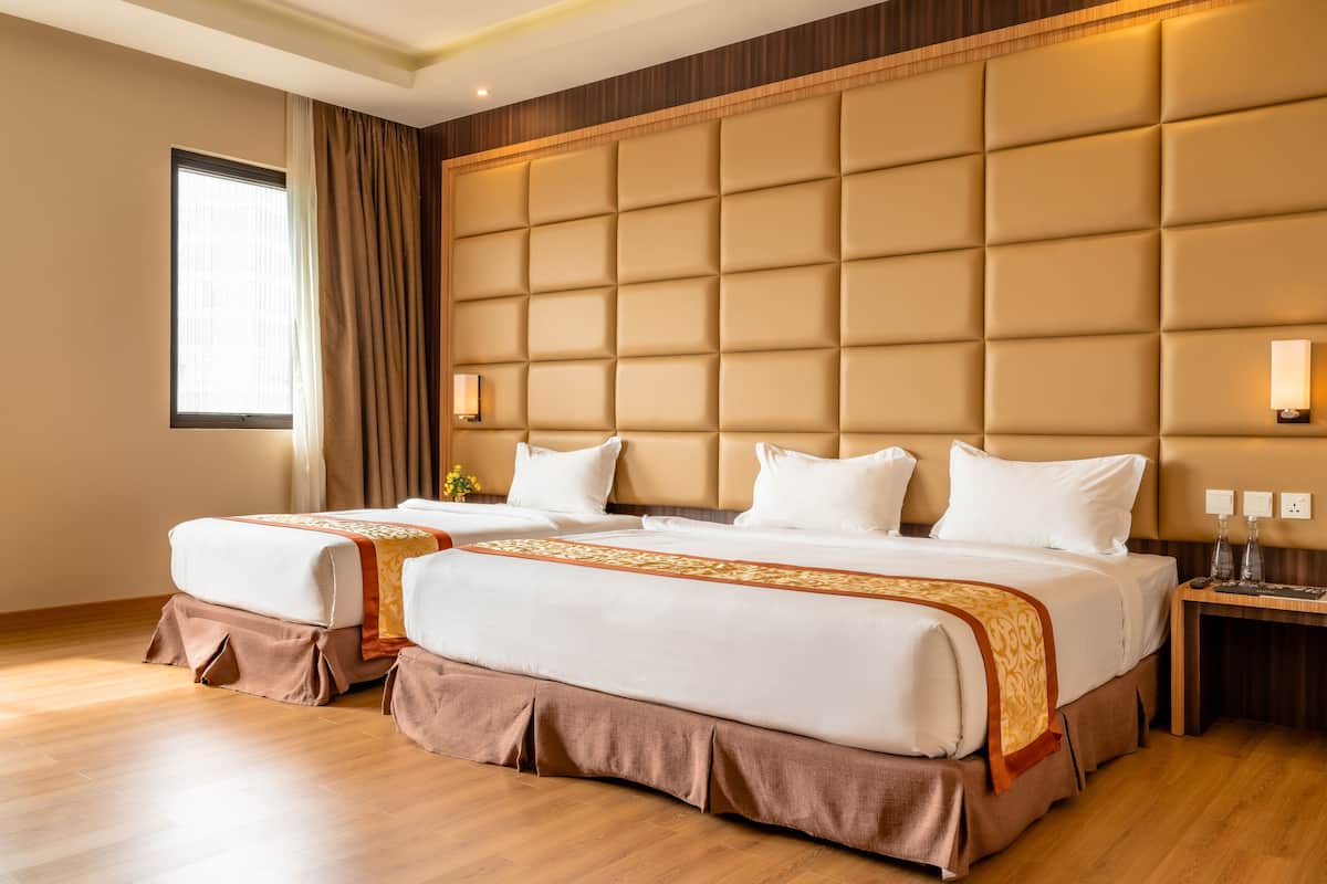 Quarto de hotel com duas camas, uma cabeceira grande e acolchoada, piso de madeira e uma janela com cortinas.