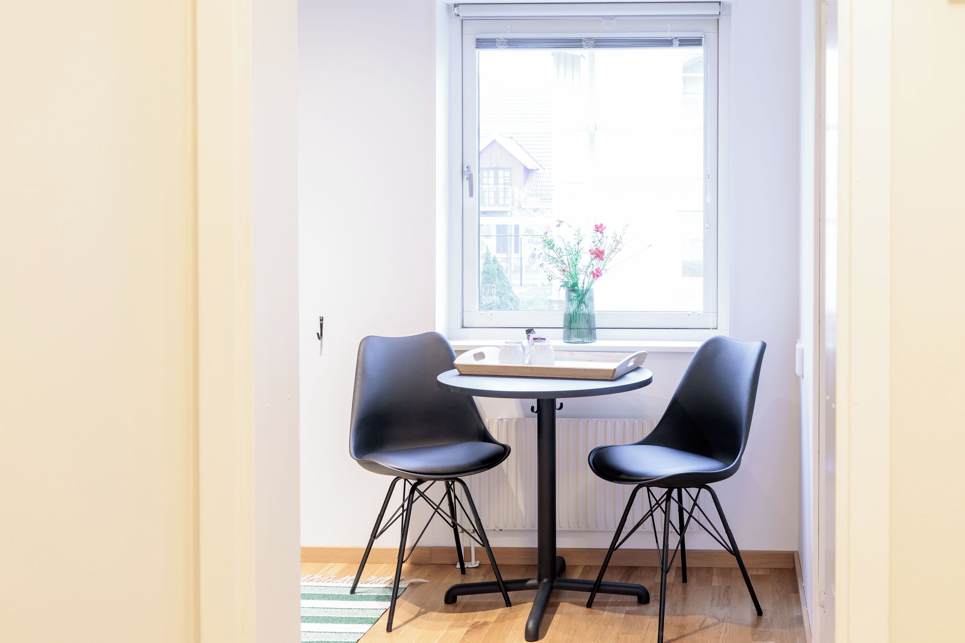Um quarto pequeno com uma mesa redonda e duas cadeiras pretas, uma janela com vista para casas, e um vaso com flores sobre a mesa.