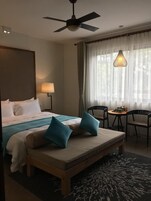 Quarto de hotel com cama, um banco, duas cadeiras, uma mesa, um ventilador de teto, abajur e janela com cortinas.