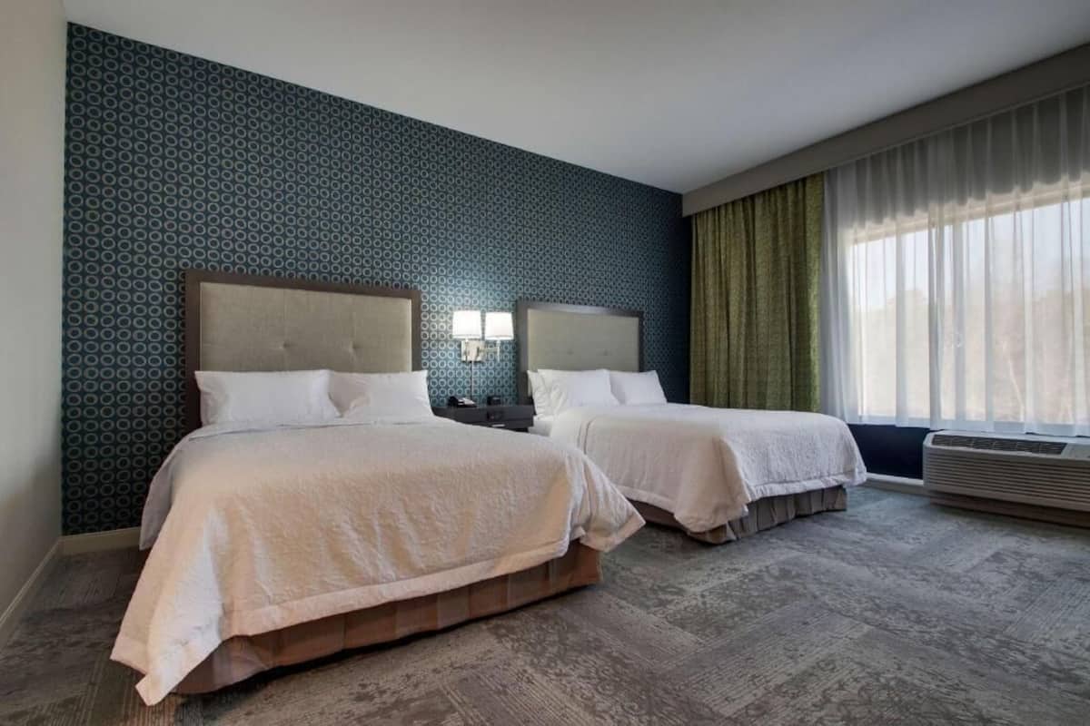 Quarto de hotel com duas camas, parede estampada, janela grande com cortinas e unidade de ar condicionado visível.