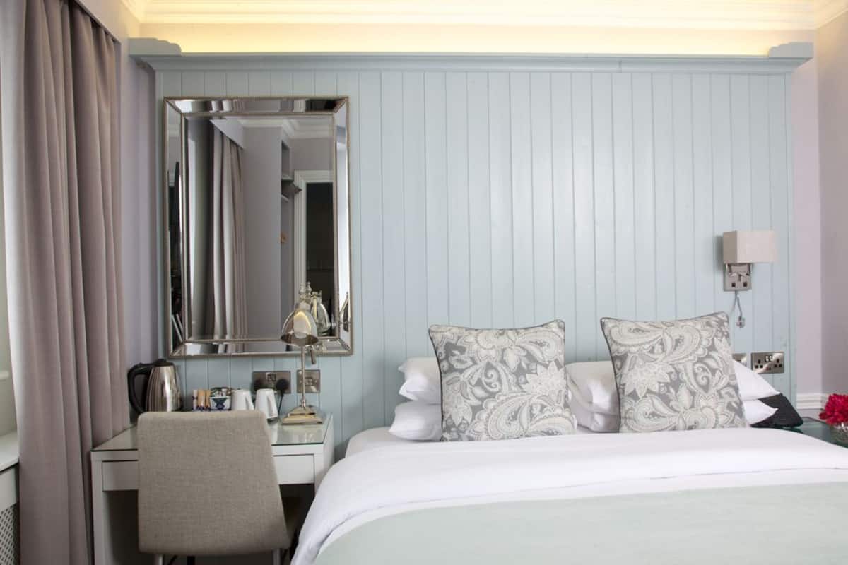 Quarto de hotel com uma cama, escrivaninha, cadeira, espelho e abajur.