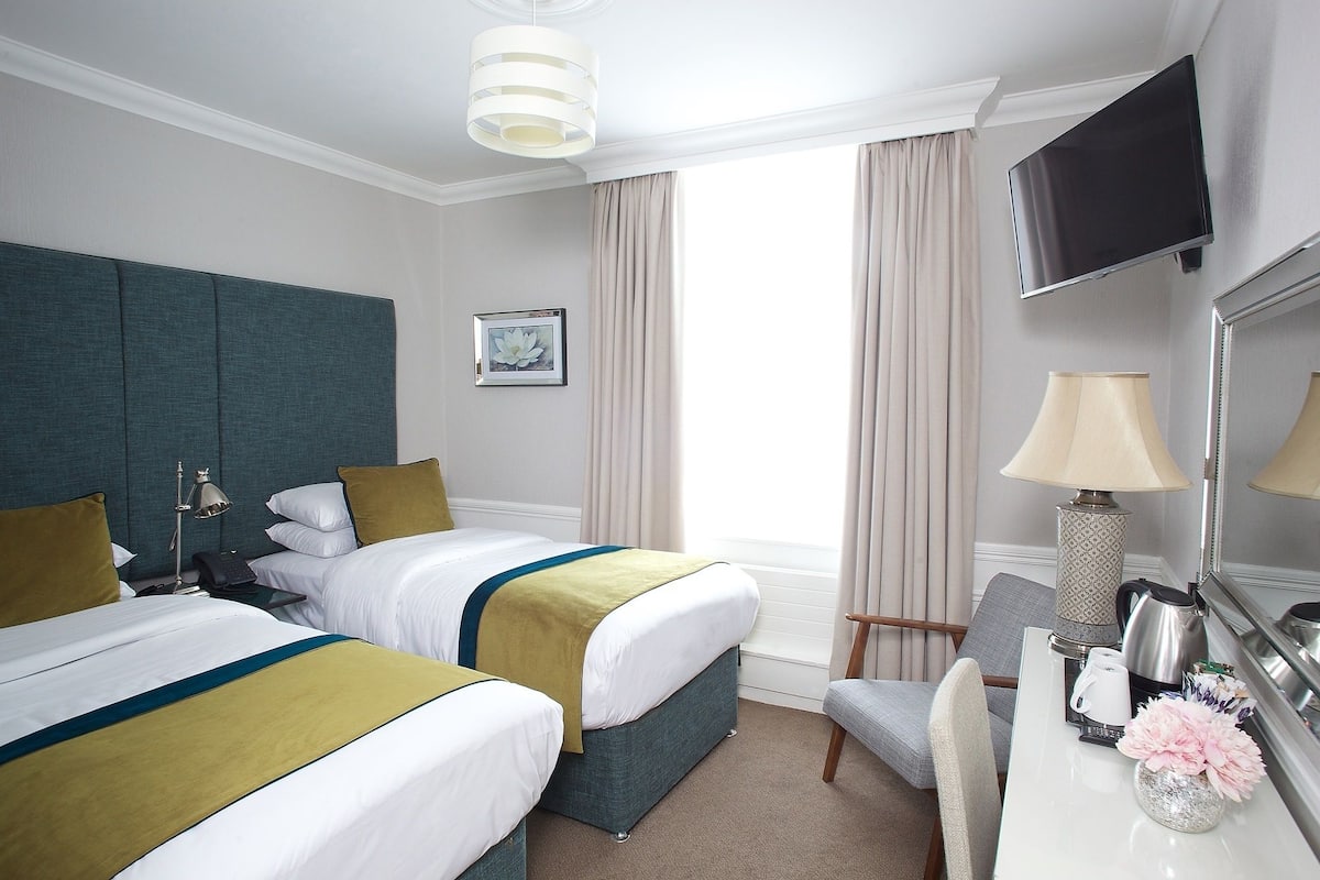Quarto de hotel com duas camas, uma escrivaninha, televisão e janela com cortinas.