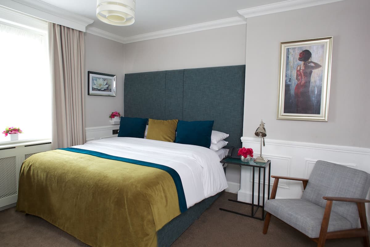 Quarto de hotel com uma cama grande, uma mesinha de cabeceira, uma cadeira, um quadro na parede e uma janela com cortinas.
