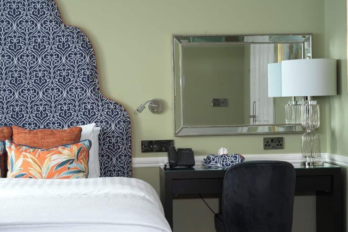 Quarto de hotel com cama, escrivaninha com telefone, cadeira, espelho e abajur.