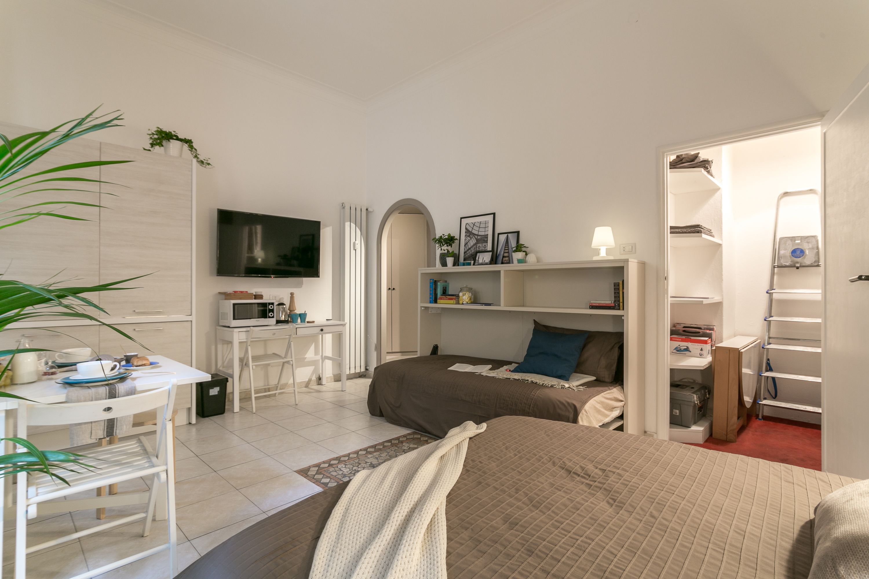 Quarto moderno com cama, mesa de cabeceira, televisão e pequena área de trabalho.