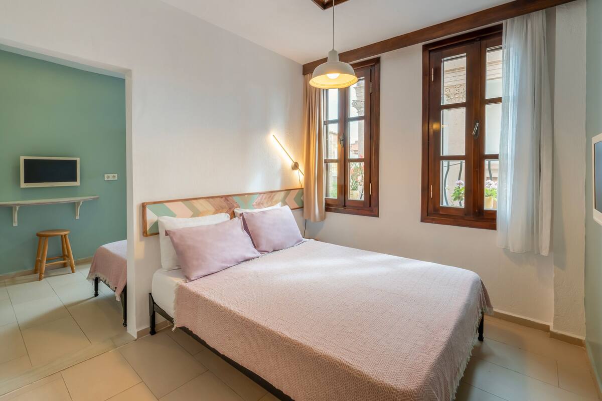 Quarto com cama, mesa de cabeceira, televisão em uma estante e janela com cortinas.