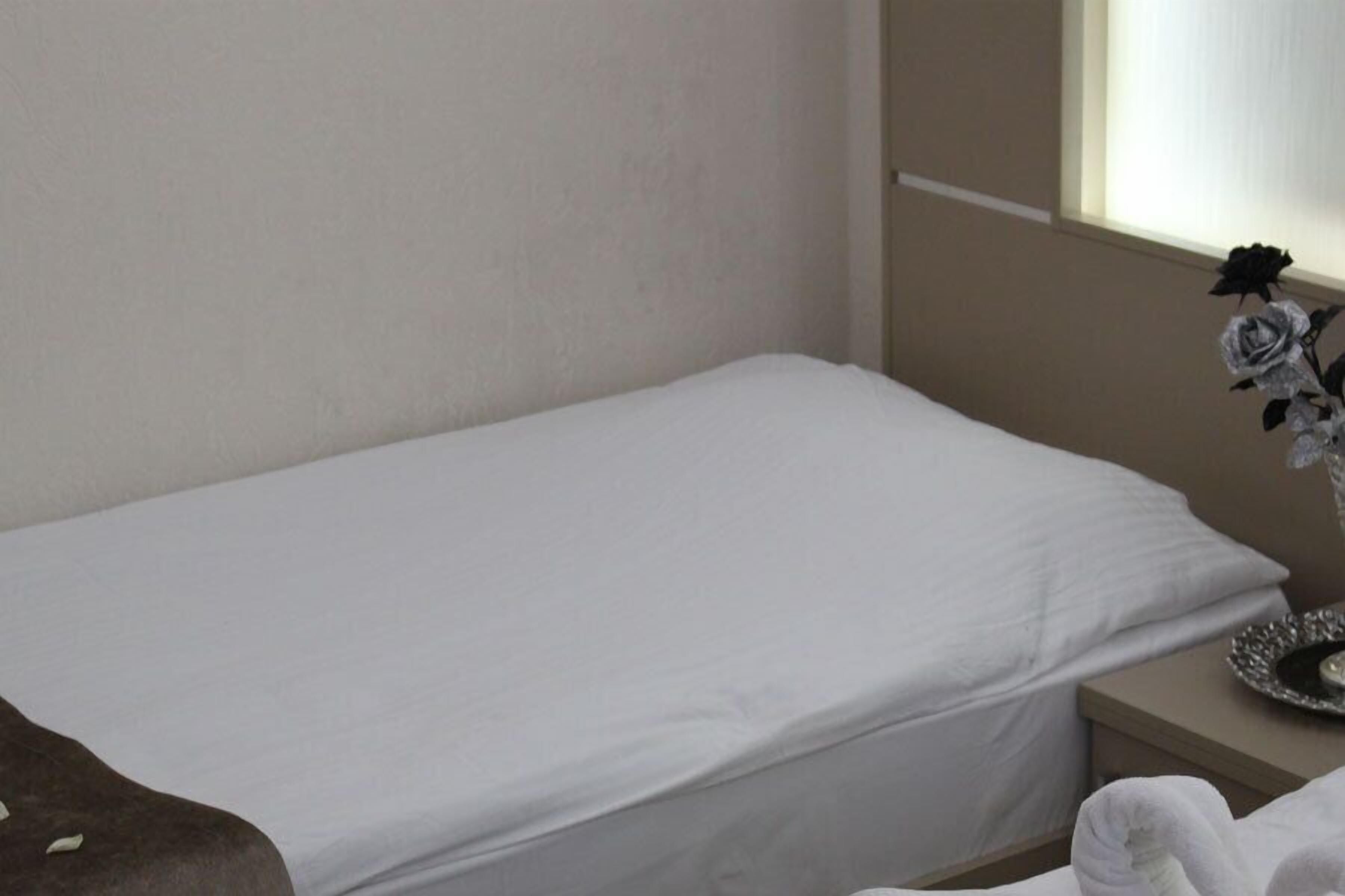 Uma cama de solteiro com lençol branco, uma mesinha de cabeceira com um prato decorativo e um vaso com flores na mesa de apoio.