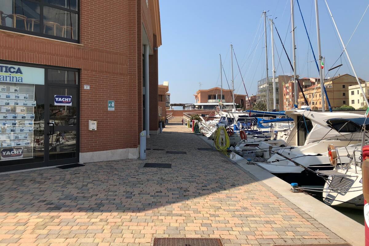 Uma marina com barcos atracados, um edifício de tijolos com placa indicando uma corretora de iates, e um céu claro.
