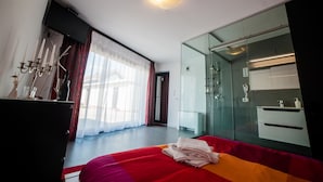 Quarto de hotel moderno com cama, box de vidro, TV de tela plana e uma grande janela com cortinas translúcidas.
