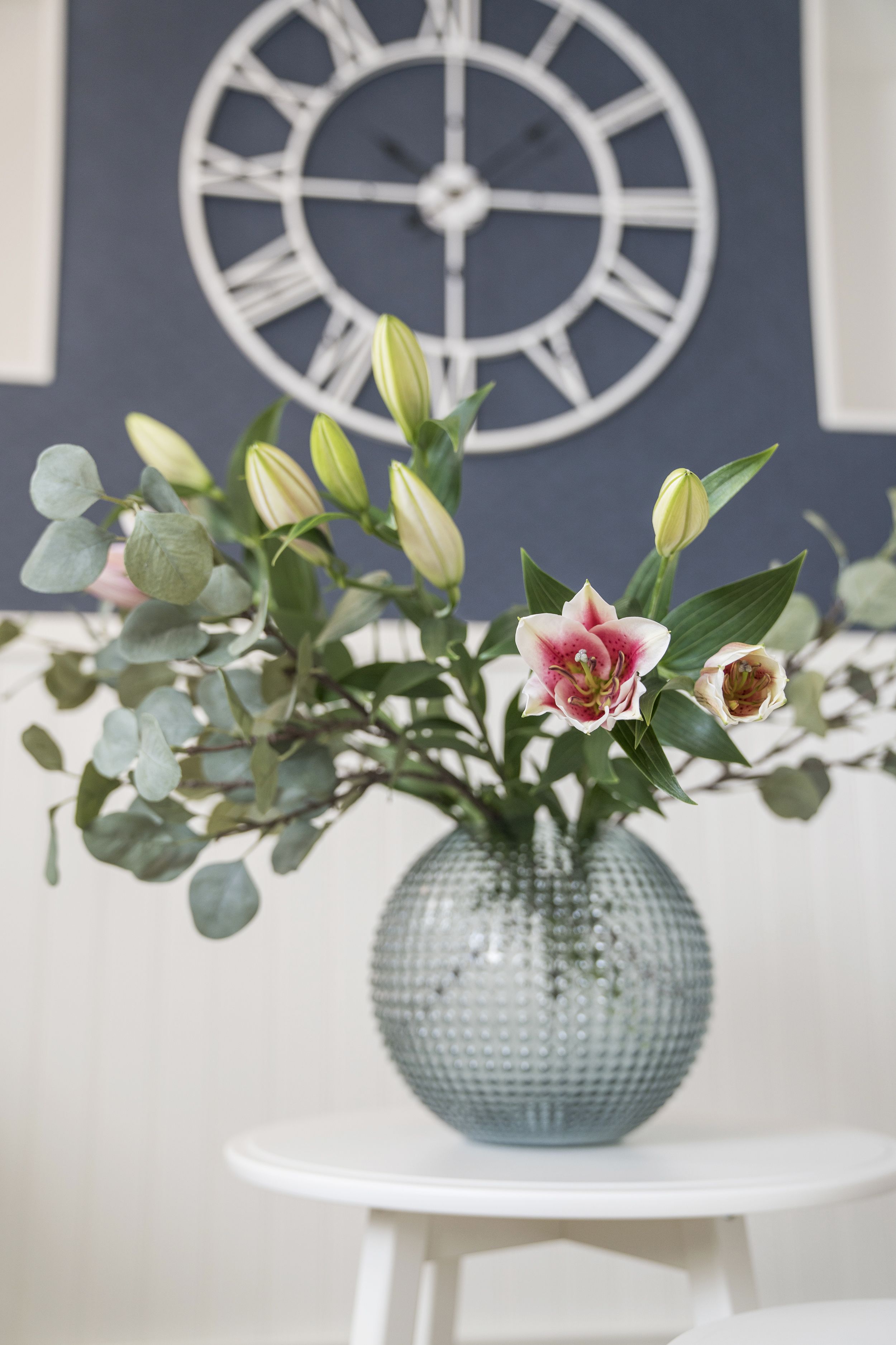 Um vaso com flores e botões sobre uma mesa branca, em frente a uma parede com um relógio grande.