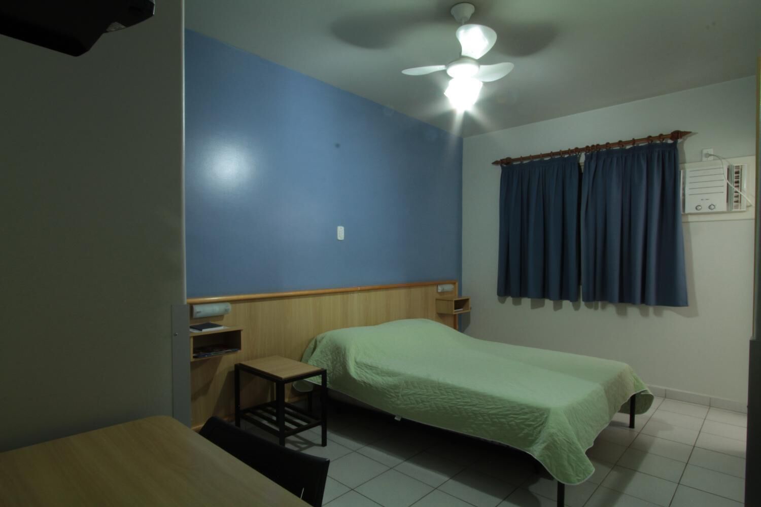 Quarto pequeno e modesto com uma cama, uma escrivaninha, uma cadeira, um ventilador de teto e uma janela com cortinas.