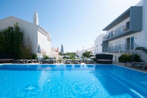 Piscina com espreguiçadeiras e guarda-sóis, cercada por edifícios com varandas e um céu claro.