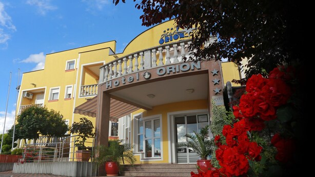 Entrada de hotel com fachada amarela, uma varanda e flores vermelhas em primeiro plano.