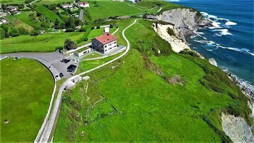 Vista aérea de uma propriedade costeira com uma casa grande, uma estrada sinuosa e um penhasco com vista para o oceano.