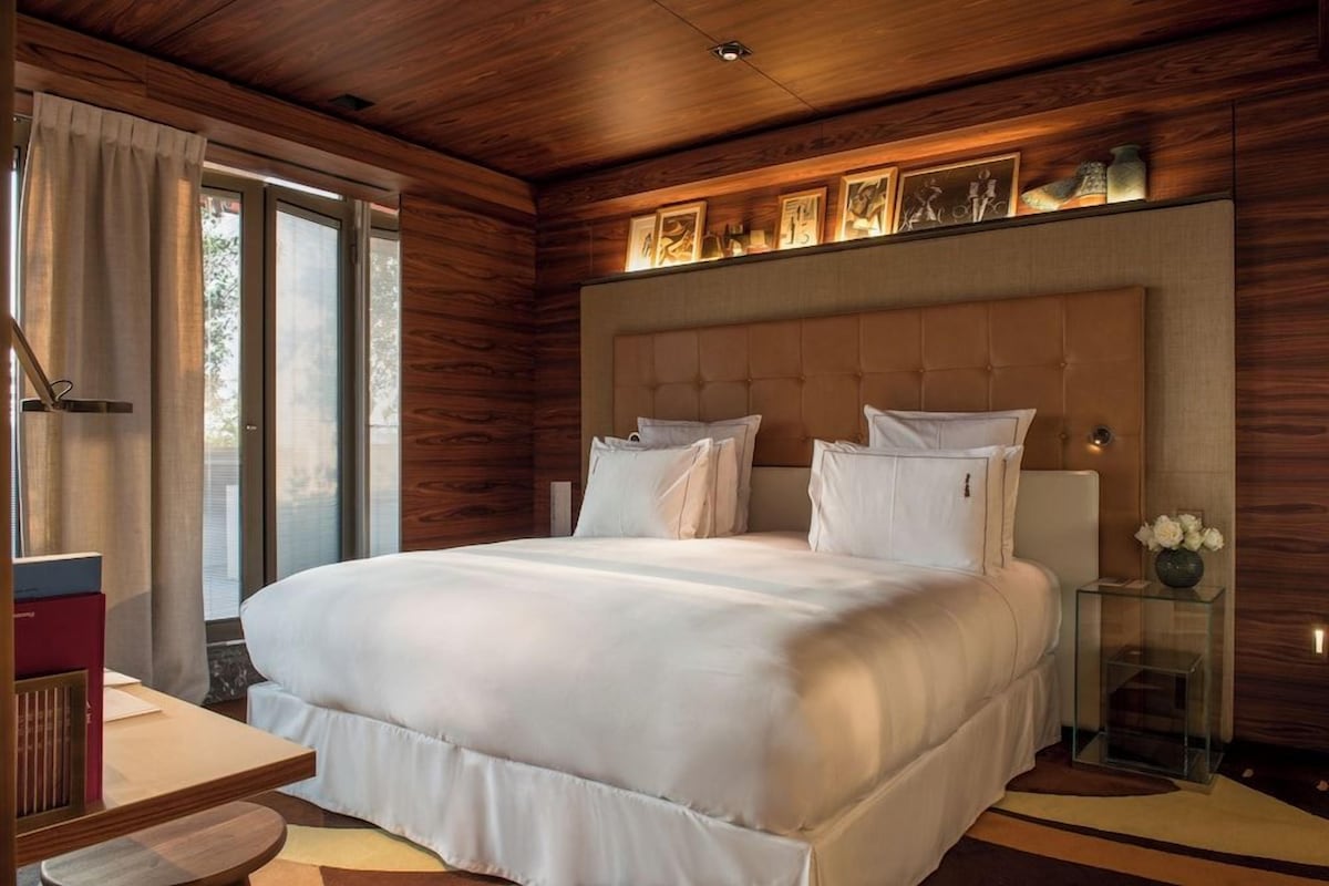 Uma cama grande com cabeceira acolchoada, mesa de cabeceira com um vaso de flores e vista para o exterior através de uma porta deslizante.