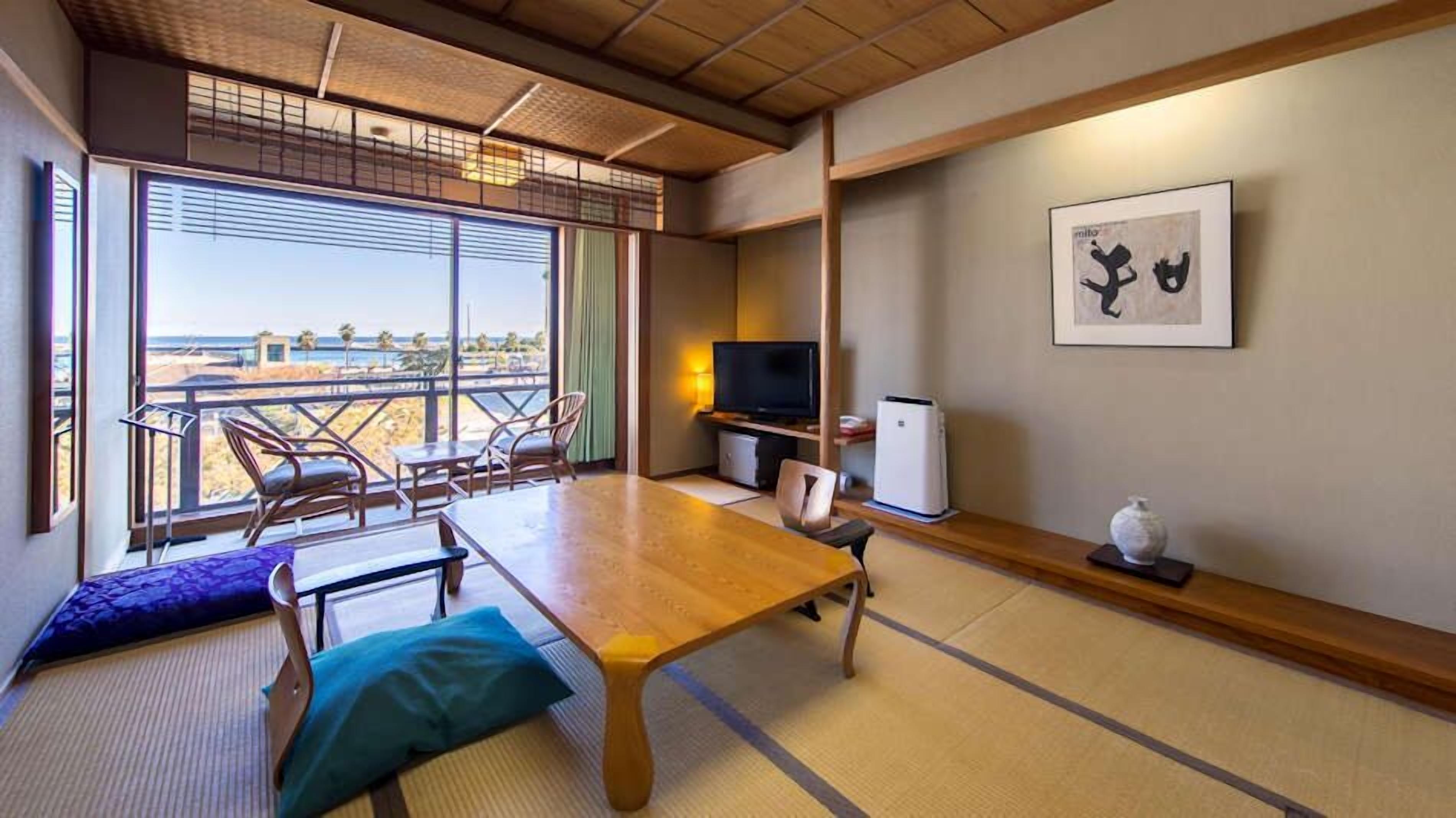 Quarto com piso de tatame, uma mesa baixa, cadeiras, uma televisão, um pequeno frigorífico e um quadro na parede.