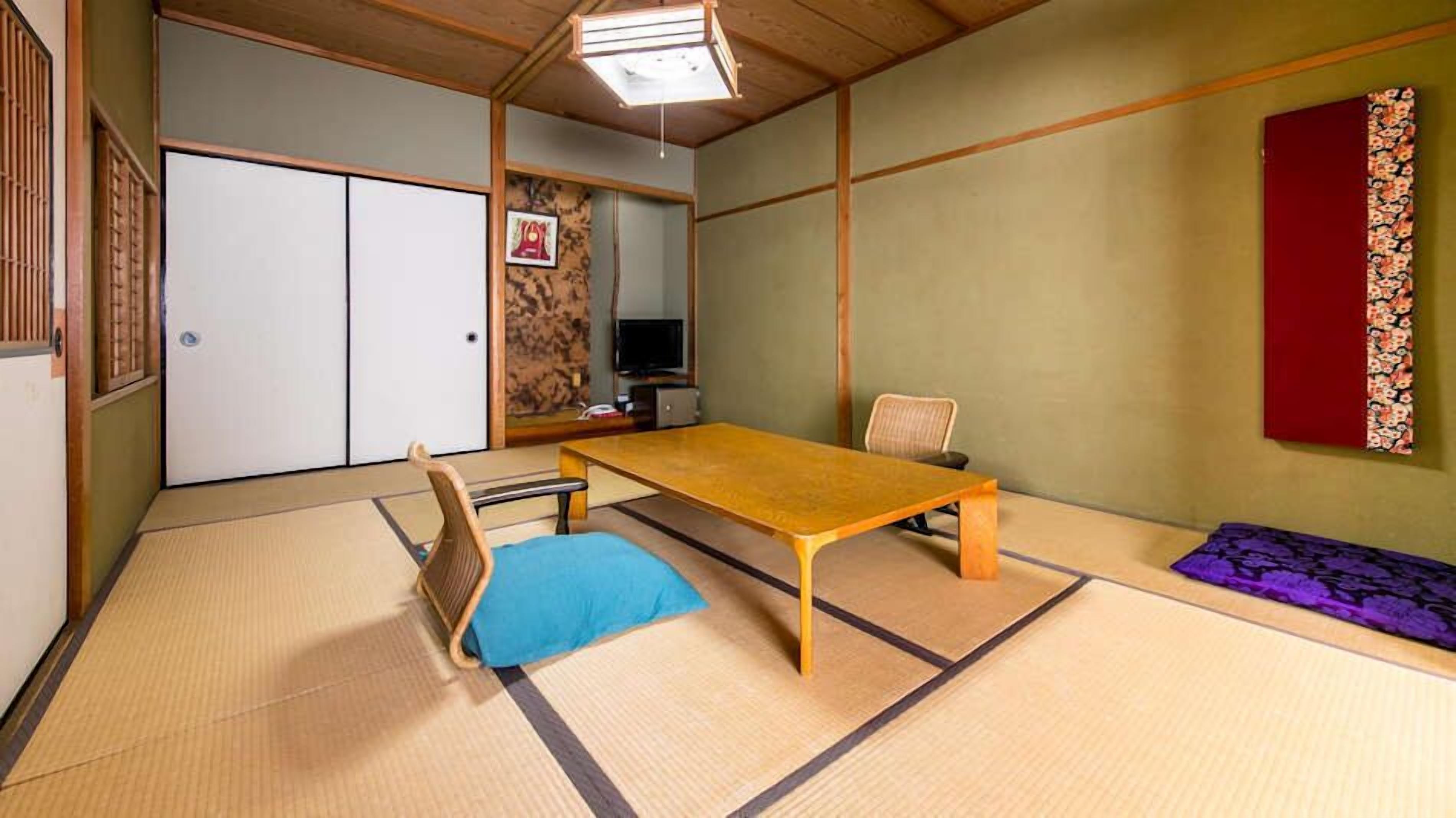 Um quarto com tatames, uma mesa baixa, uma cadeira, uma televisão e uma cortina vermelha.